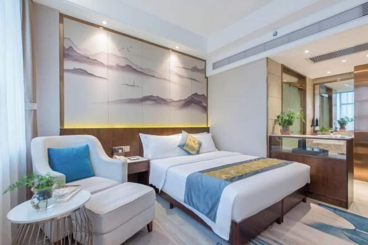 Quarto de hotel com uma cama, uma mesa de escritório, uma cadeira, uma mesinha com uma planta e uma pintura grande na parede.