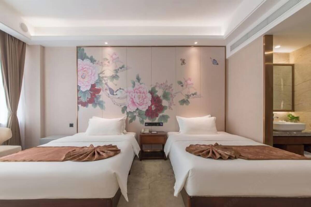 Quarto de hotel com duas camas, uma grande obra de arte floral na parede, uma mesa de cabeceira com abajur, e banheiro com espelho e pia.
