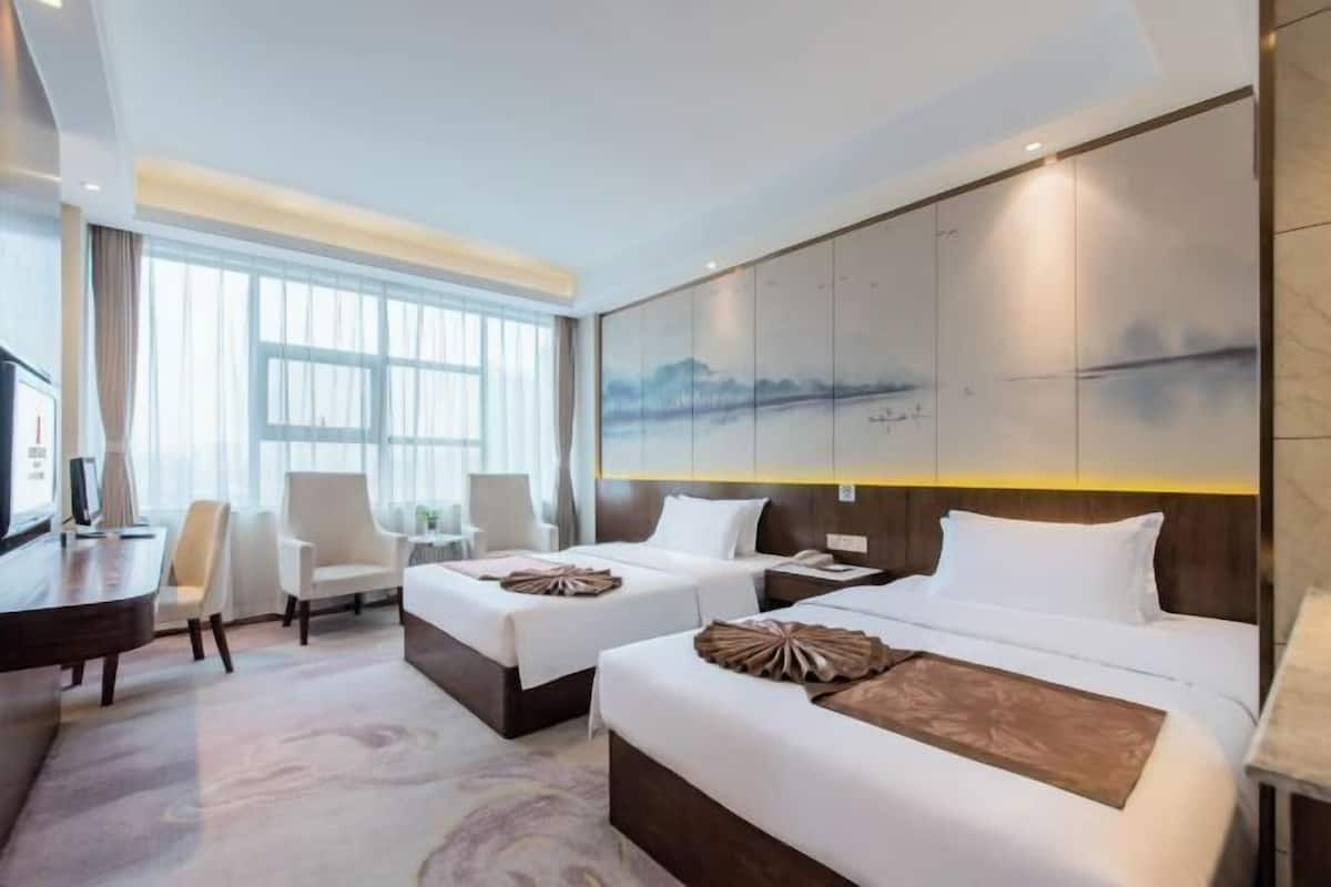 Quarto de hotel com duas camas, uma escrivaninha, cadeiras e uma janela grande.
