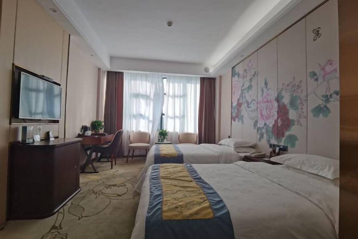 Quarto de hotel com cama, escrivaninha, televisão e uma janela grande com cortinas.