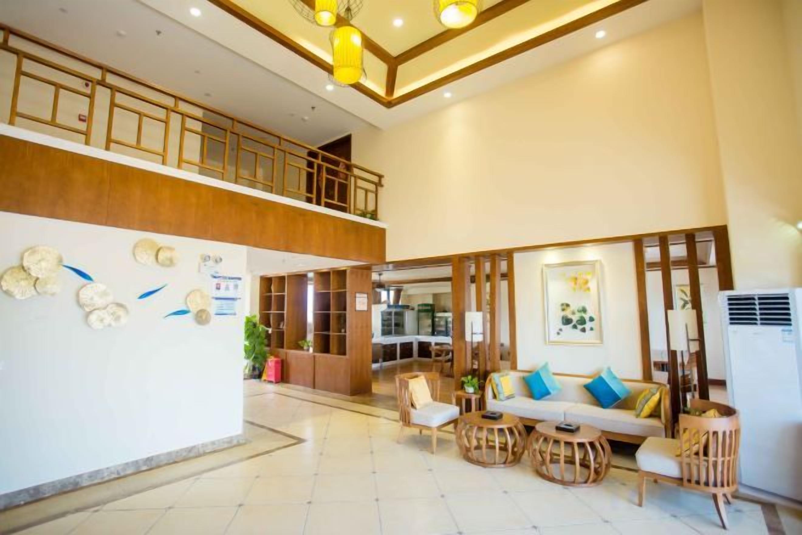 Um saguão de hotel com balcão de recepção, área de estar com cadeiras e sofás, escada e uma grande janela.