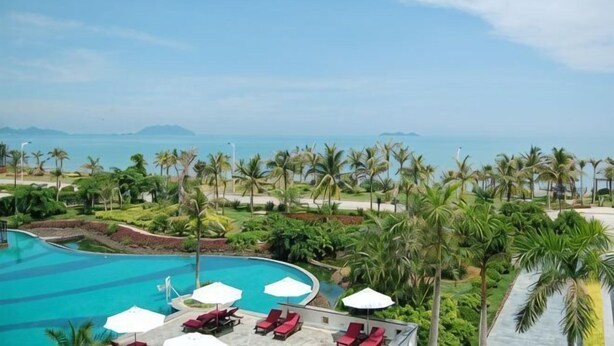 Piscina de resort com cadeiras de lounge e guarda-sóis, cercada por palmeiras e vista para o mar.