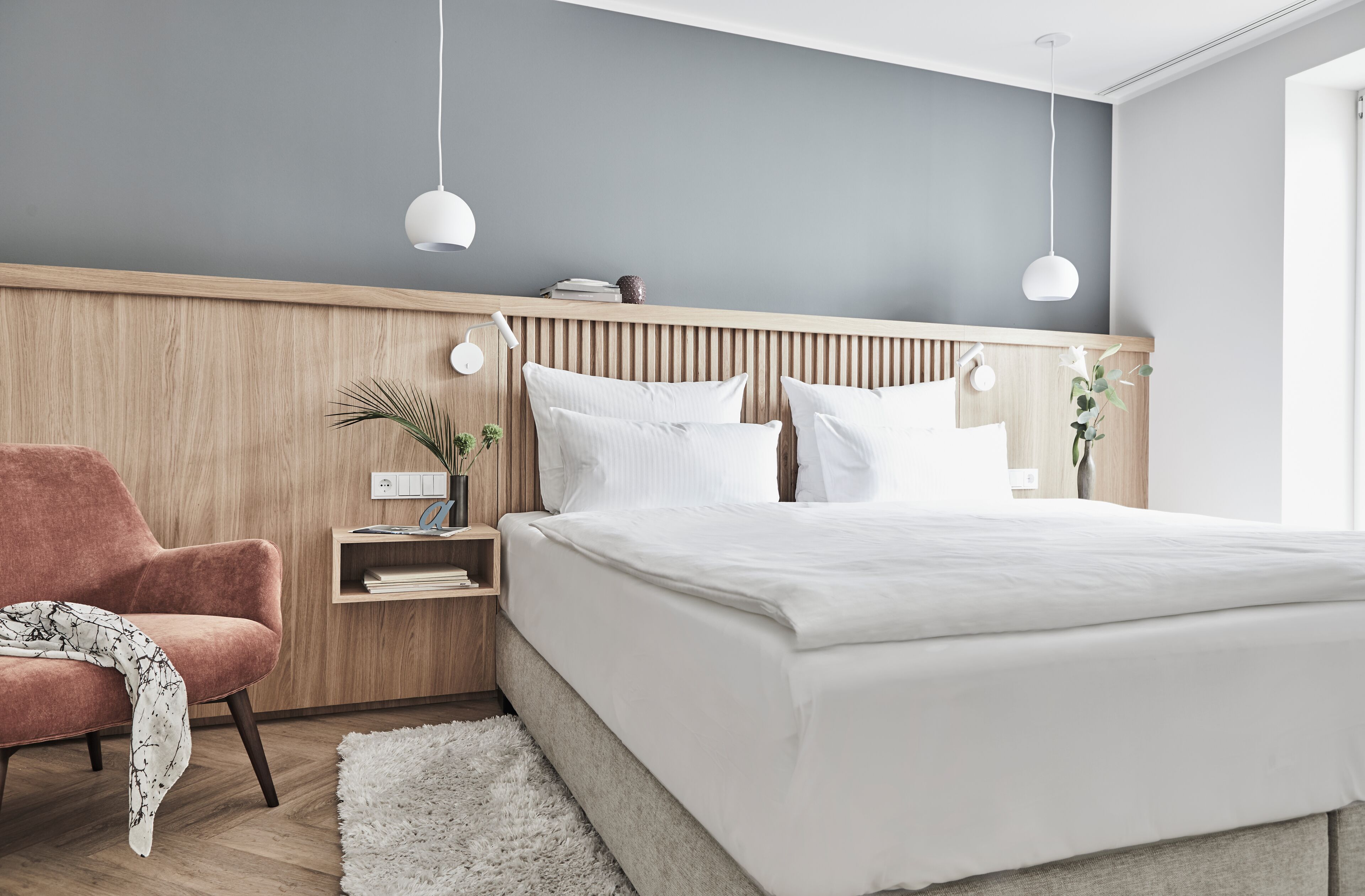 Quarto moderno com cabeceira de madeira, cama branca, poltrona rosa e tapete branco.