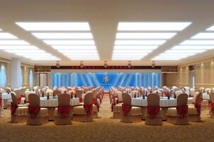 Sala de conferências com mesas e cadeiras redondas, palco com fundo azul e piso coberto de carpete.