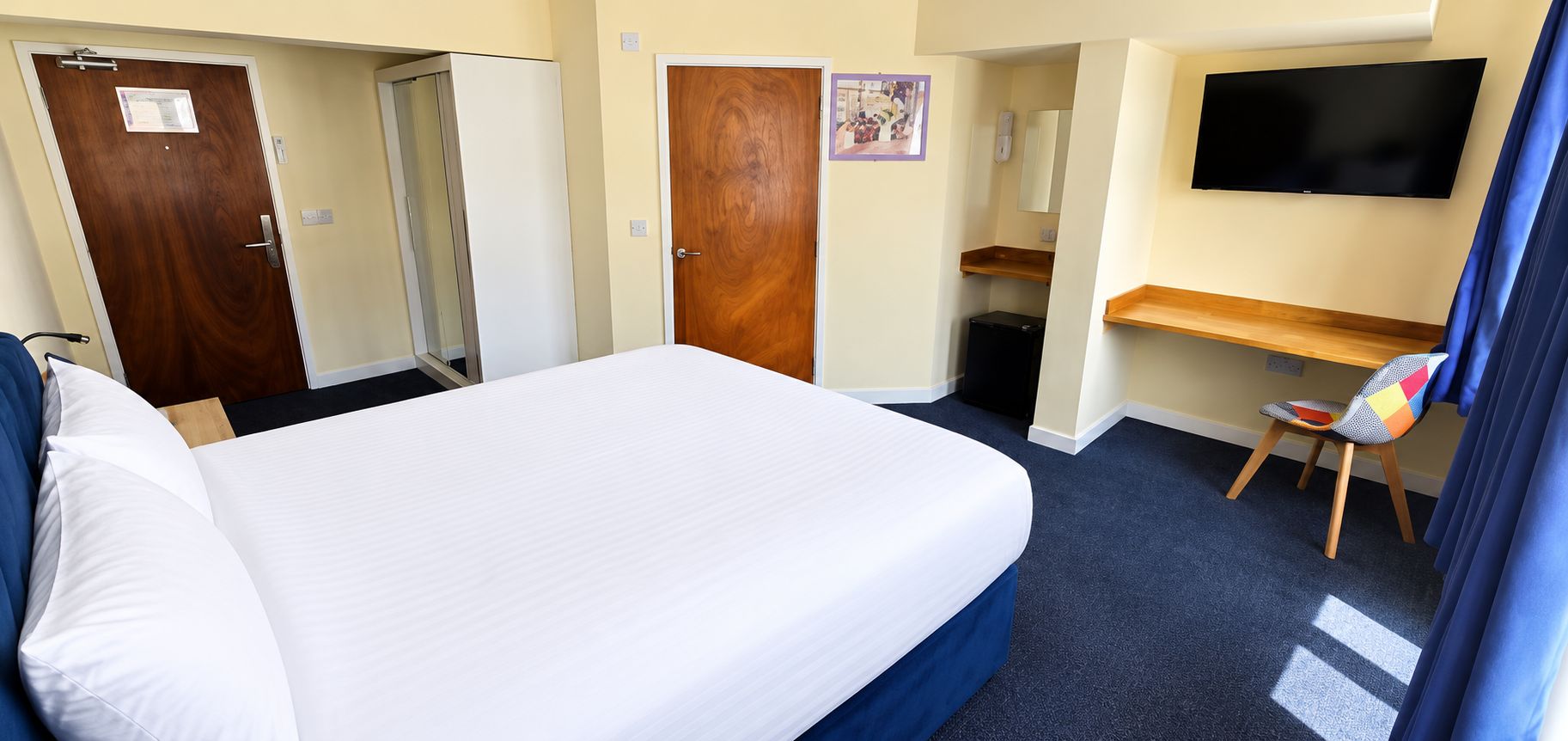 Quarto de hotel com cama, escrivaninha, cadeira, televisão e porta.