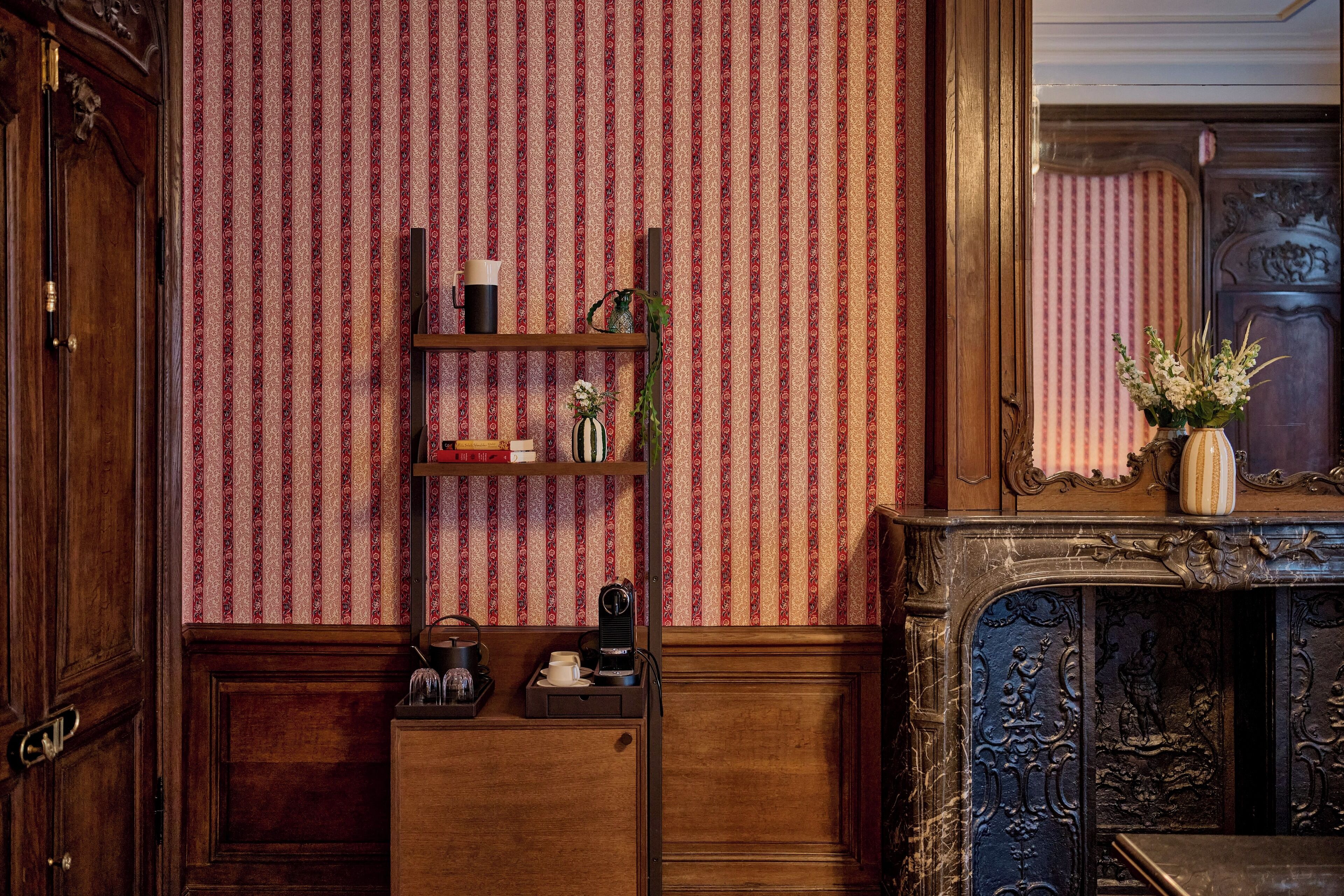 Um quarto com papel de parede estampado, uma prateleira de madeira com diversos objetos e uma lareira com detalhes entalhados.