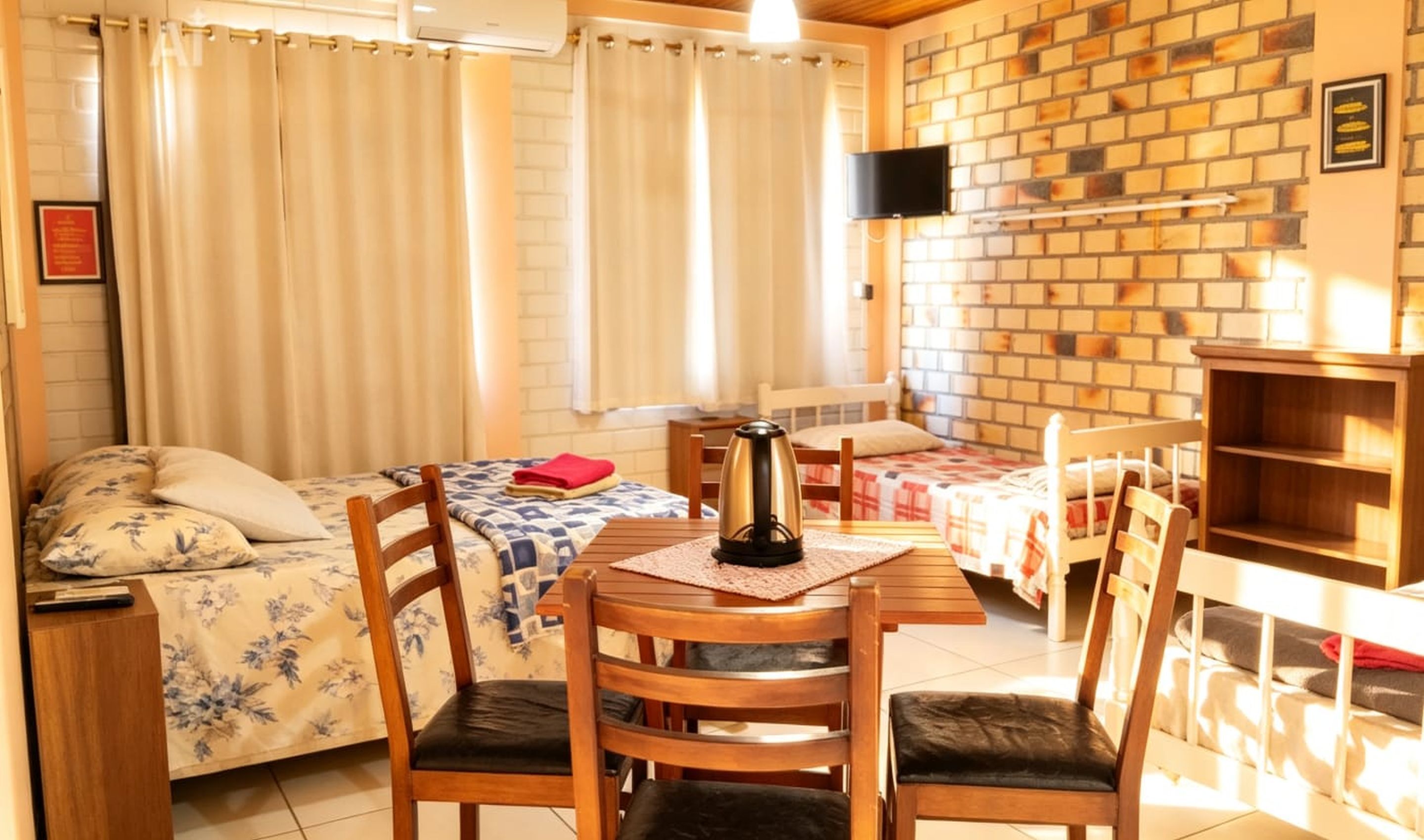 Um quarto aconchegante com duas camas, uma mesa com cadeiras, uma chaleira e uma parede de tijolos.