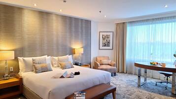 Quarto de hotel com uma cama grande, duas mesas de cabeceira, uma cadeira, uma escrivaninha com cadeira e uma janela com cortinas.