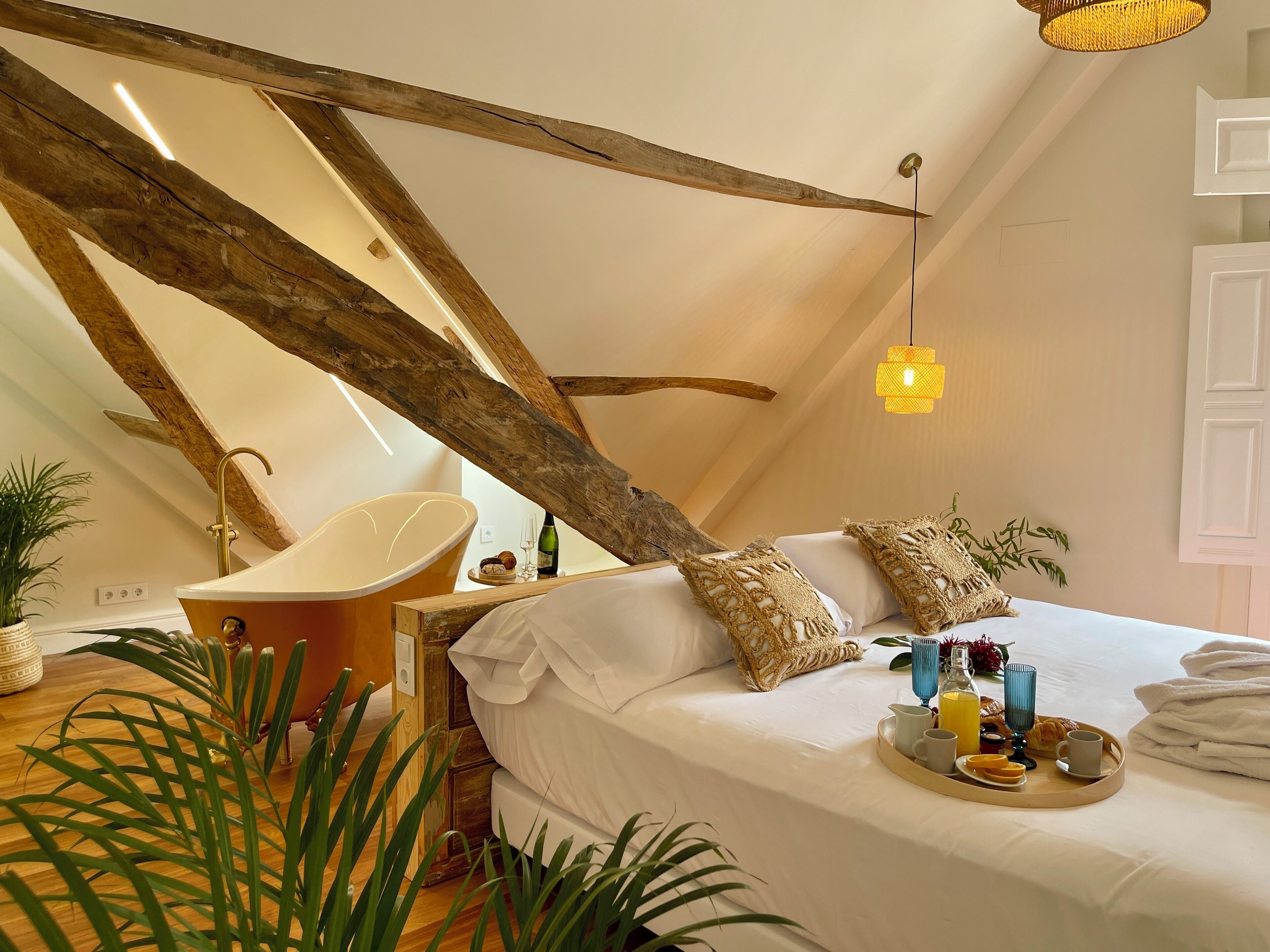Um quarto aconchegante com teto inclinado, estrutura de vigas de madeira, cama branca com almofadas decorativas, mesa de cabeceira com garrafa e copos, e bandeja com itens de café da manhã.