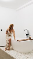 Mulher de vestido branco ao lado de uma banheira branca, com uma bandeja de madeira, cercada por pedrinhas brancas.