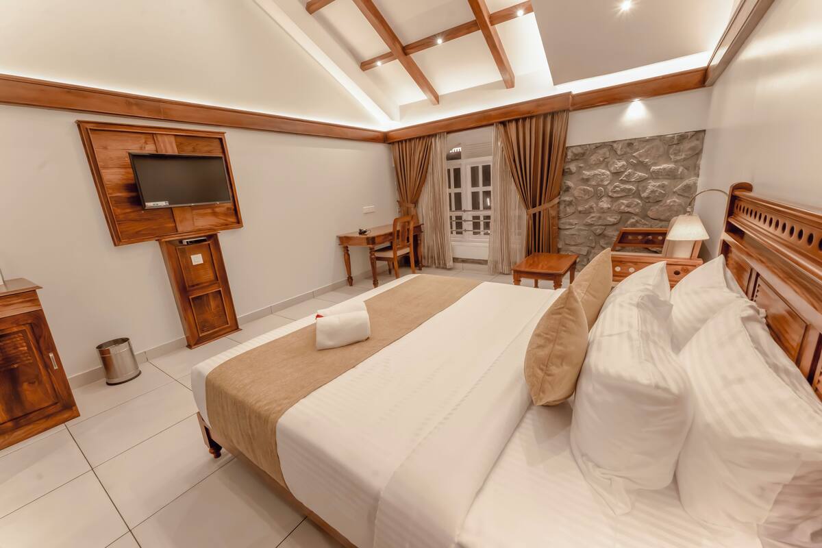 Quarto de hotel moderno com uma cama grande, mobiliário de madeira, televisão fixada na parede e uma parede de destaque em pedra.