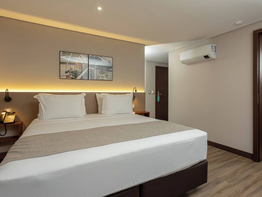 Quarto de hotel moderno com uma cama grande, abajures, telefone, ar condicionado e vista para o banheiro.
