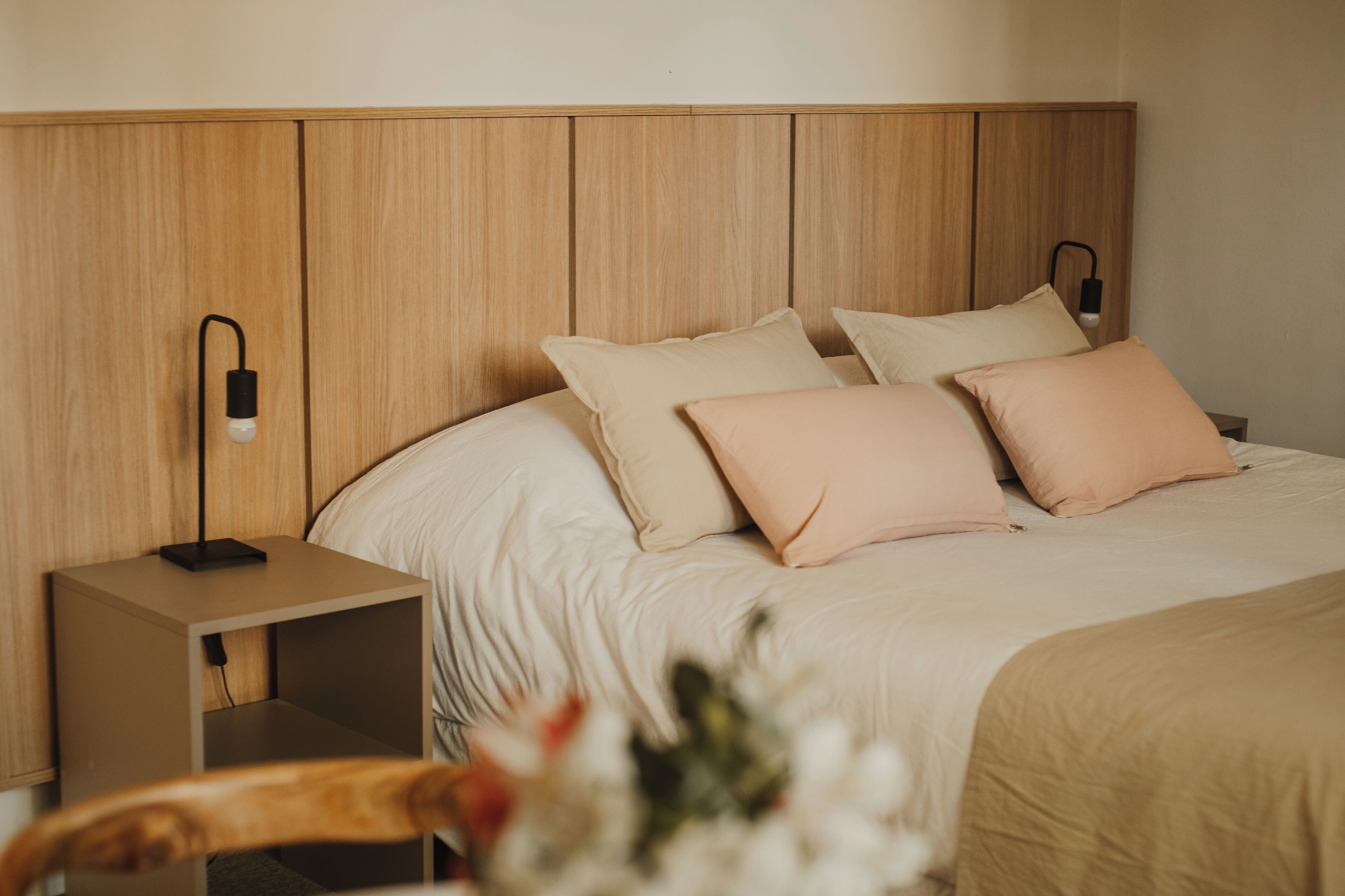 Uma cama bem arrumada com cabeceira de madeira, uma mesa de cabeceira com abajur e um criado-mudo pequeno.