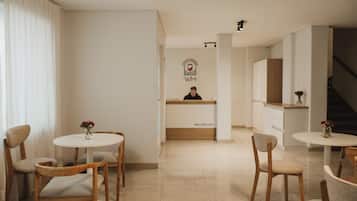Uma área de recepção de hotel com balcão de recepcionista, duas mesas redondas com cadeiras e uma escada ao fundo.
