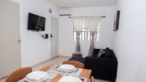 Uma sala de jantar com mesa posta para quatro, um sofá preto, uma televisão fixada na parede e um aparelho de ar condicionado.