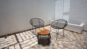 Pequena área de pátio fechada com chão ladrilhado, duas cadeiras de arame preto, uma mesa de madeira e uma parede branca com janela projetando sombras.