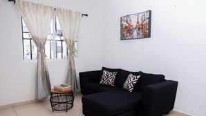 Um sofá preto com almofadas estampadas, uma mesa de apoio redonda pequena, uma janela com cortinas e um quadro de uma cena urbana.