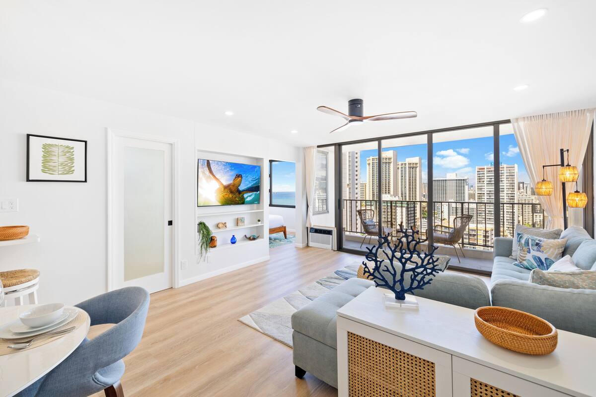 Uma sala de estar moderna com vista para a cidade, uma televisão de tela plana e um espaço de jantar com mesa branca e cadeiras azuis.