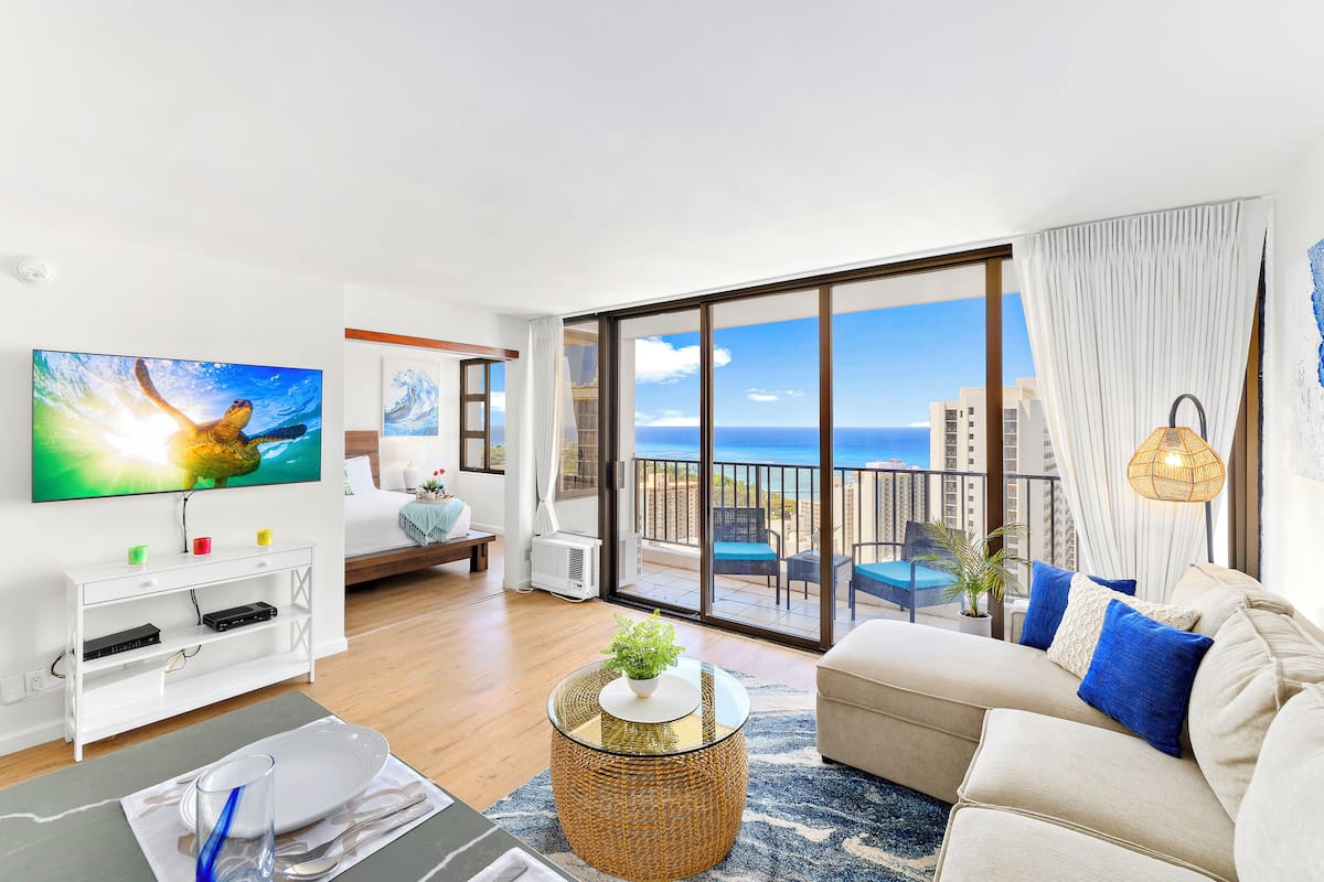 Uma sala de estar moderna com uma televisão grande, um sofá, uma mesa de centro e uma varanda com vista para o mar.