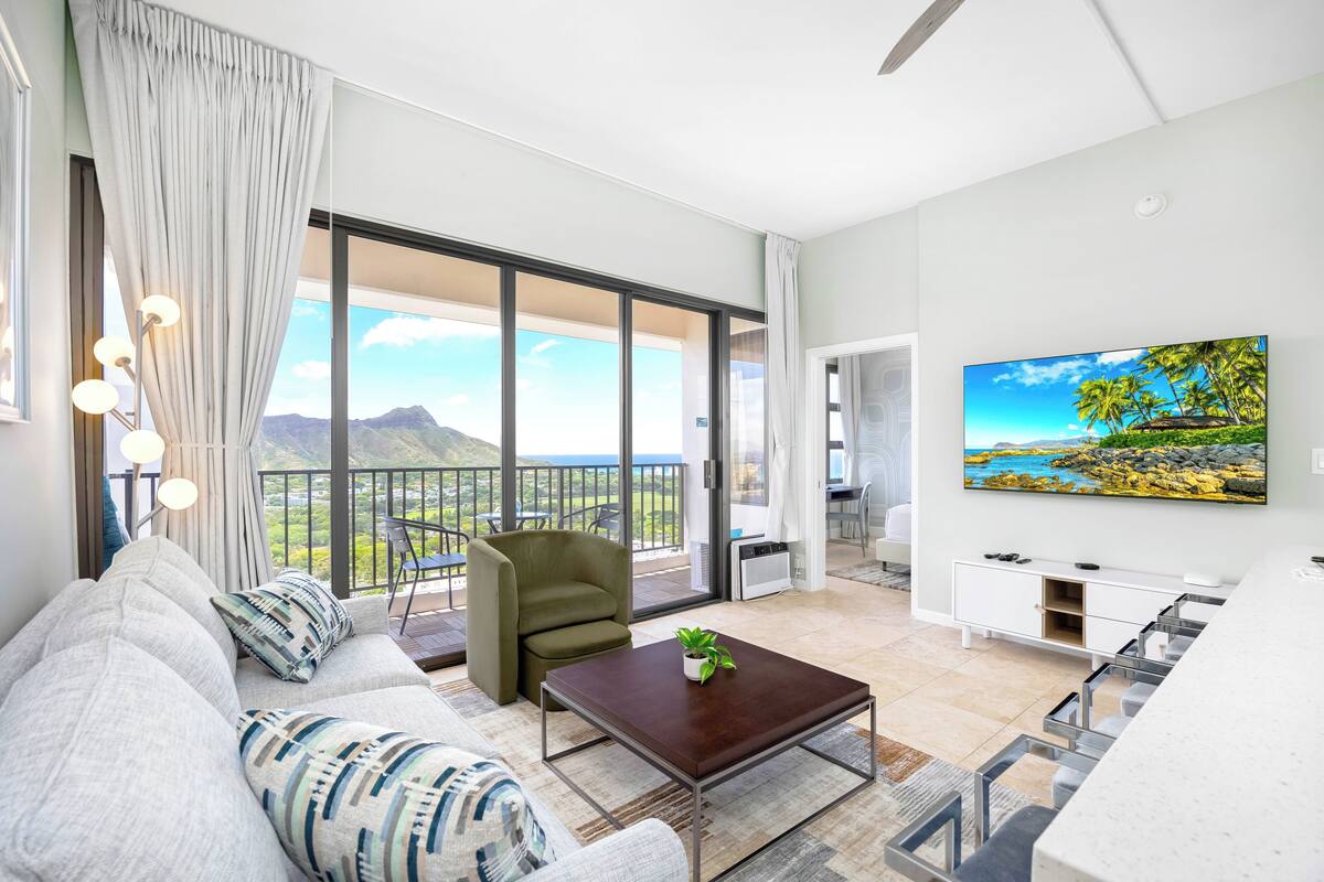 Uma sala de estar moderna com um sofá, uma mesa de centro e uma televisão. Há uma varanda com vista para montanhas e um espaço de jantar com mesa e cadeiras.