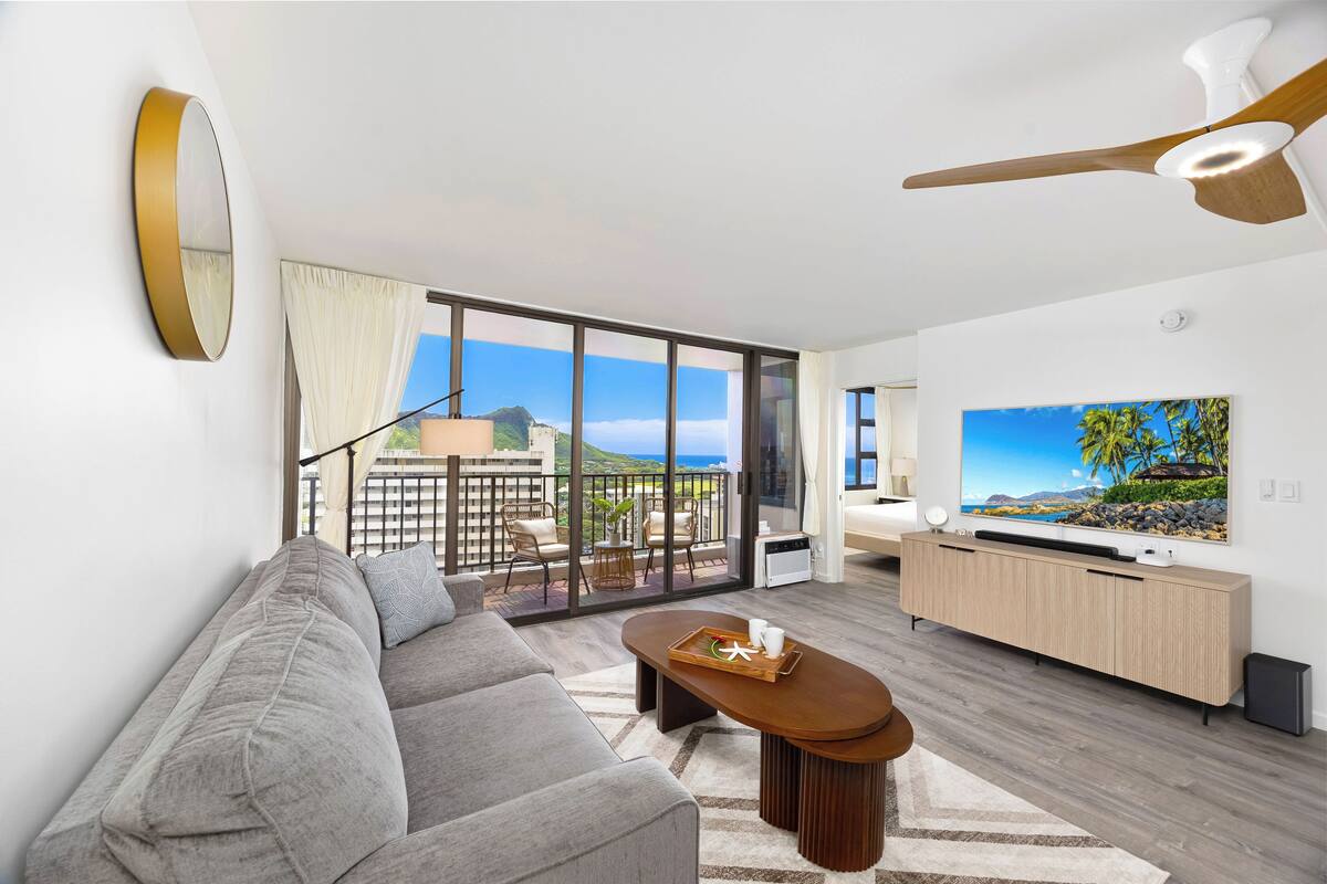 Uma sala de estar moderna com um sofá cinza, uma mesa de centro de madeira, uma TV em um móvel de madeira e uma varanda com vista para o oceano.