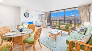 Ampla sala de estar com mesa de jantar posta, cadeiras estofadas em verde, mesa de centro de vidro e sofá com almofadas decorativas. Uma janela grande oferece vista para uma paisagem montanhosa.