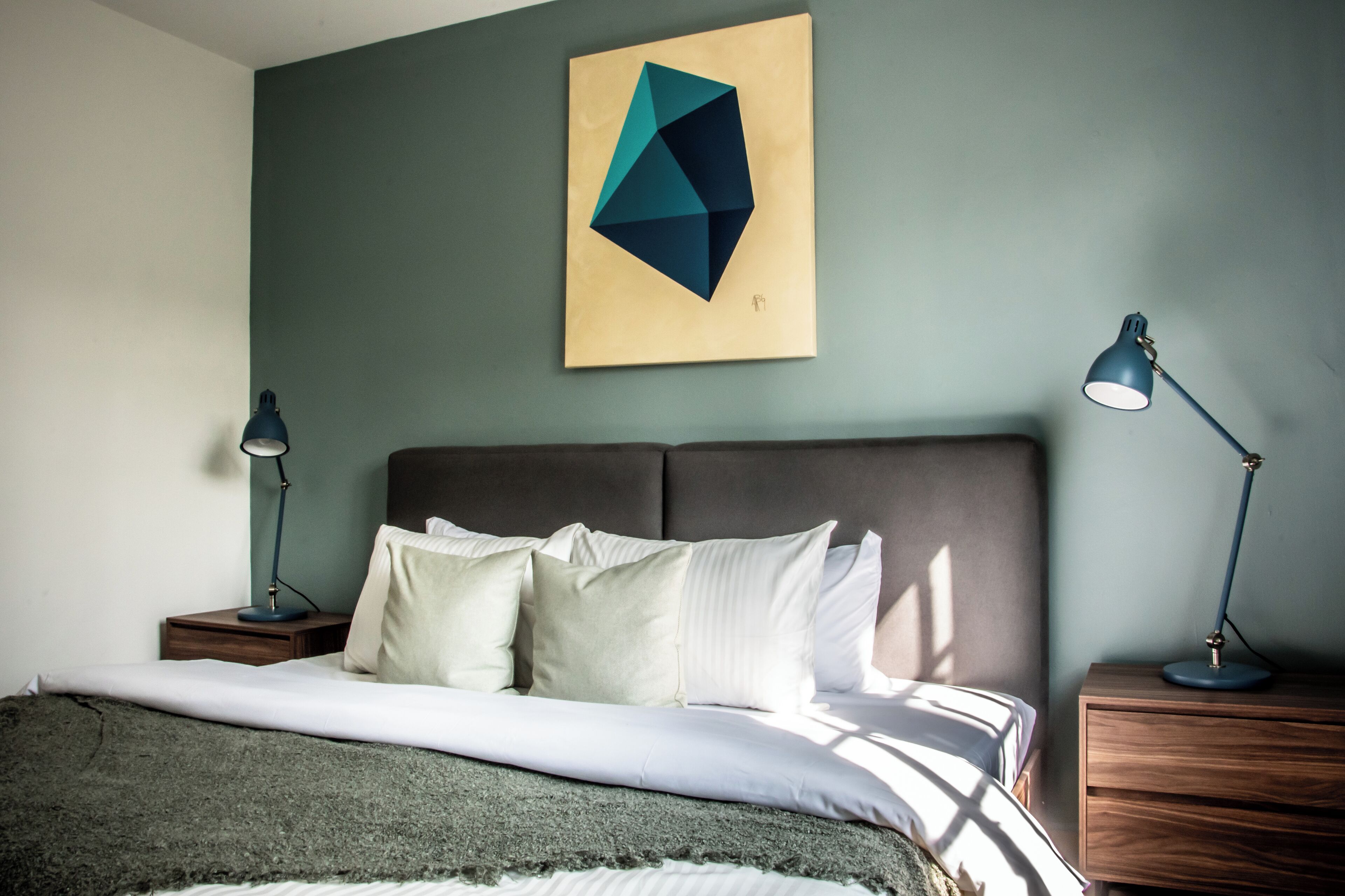 Cama bem arrumada com manta verde, duas luminárias de cabeceira e um quadro geométrico na parede.