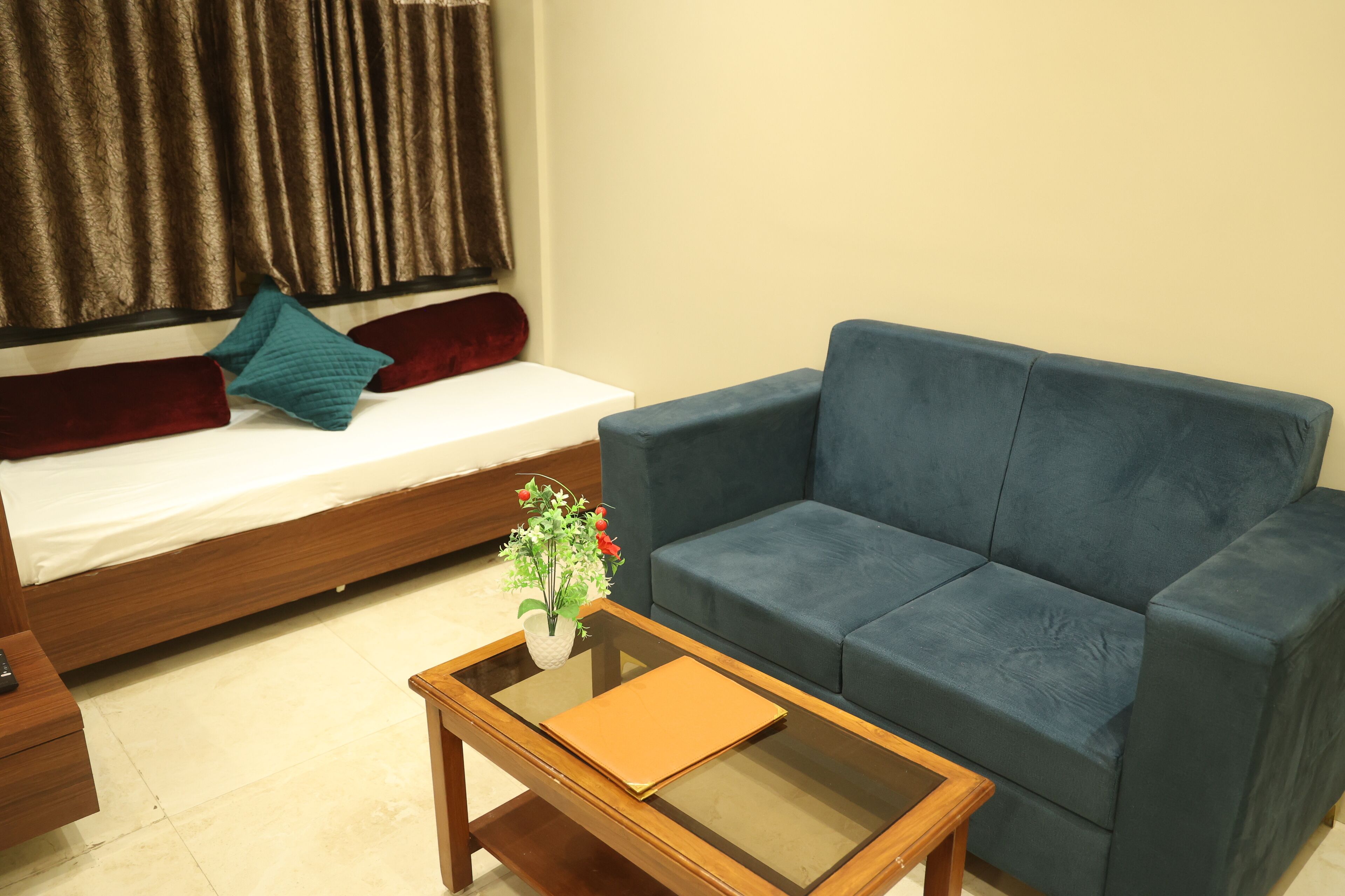 Quarto com sofá azul, mesa de centro de madeira, pequena mesa branca e cama com travesseiro vermelho e verde.