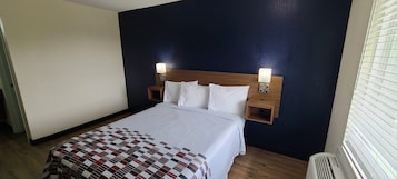 Quarto de hotel com uma cama, duas luminárias de cabeceira, um painel de cabeceira de madeira e uma janela com persianas.