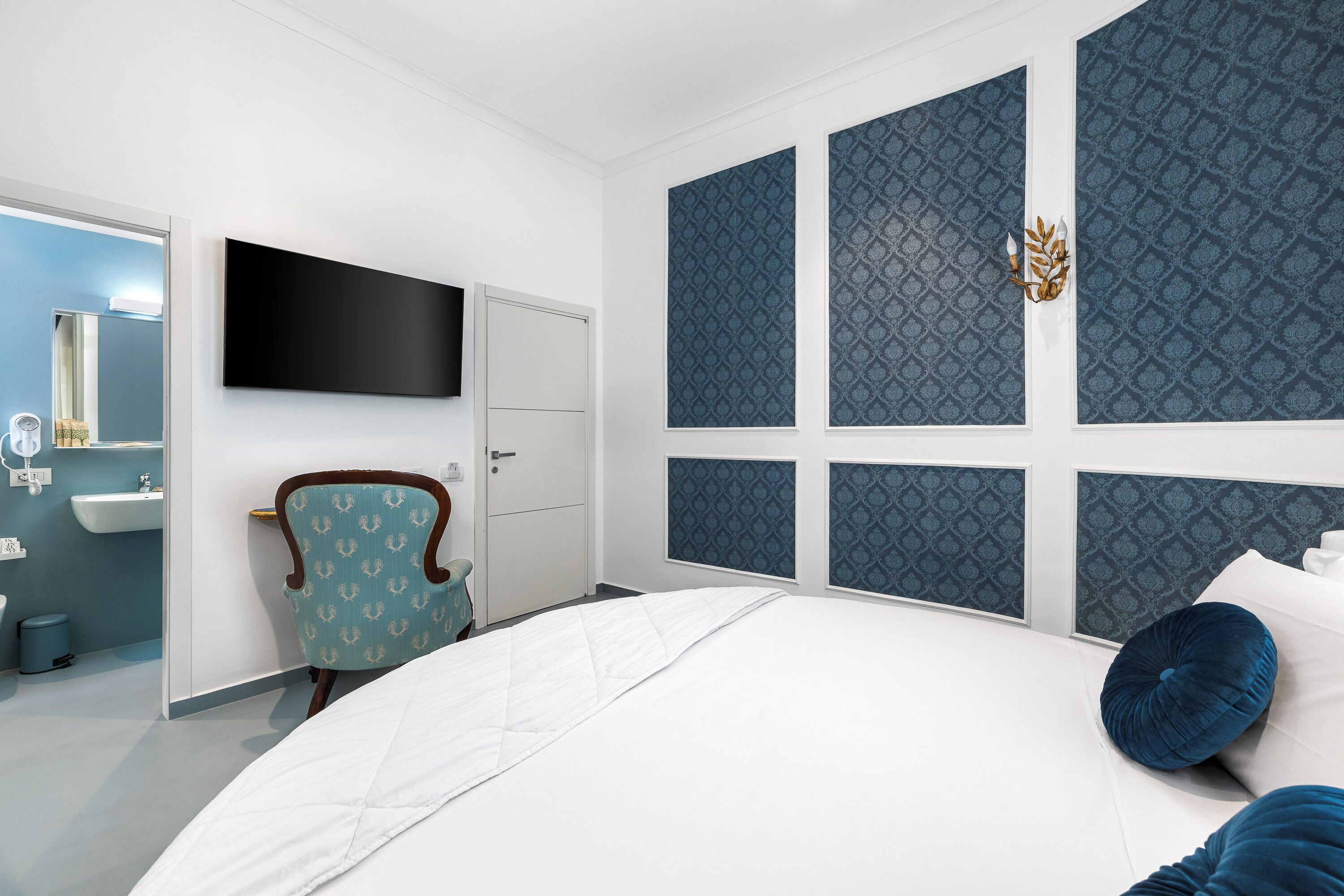 Quarto com cama, uma cadeira, TV e banheiro visível por uma porta aberta.