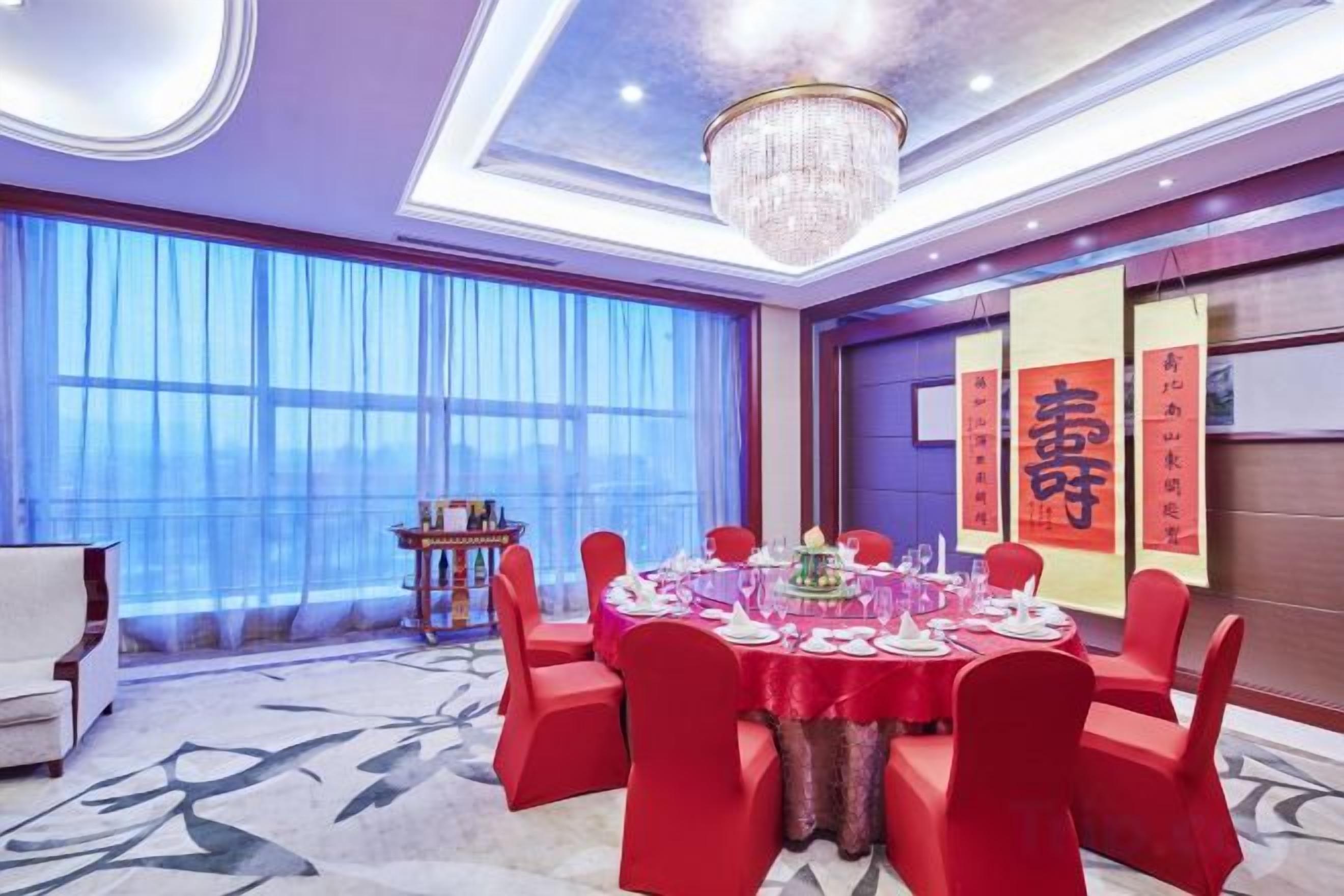 Uma sala de jantar de hotel com uma grande mesa redonda posta para refeição, cadeiras vermelhas, um candelabro e janelas amplas com cortinas.