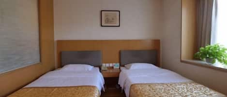Quarto de hotel com duas camas de solteiro, cabeceira de madeira, mesa de cabeceira pequena, quadro na parede e janela com planta.
