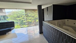 Uma casa de banho moderna com uma banheira grande, um sofá preto e vista para árvores através de uma janela ampla.
