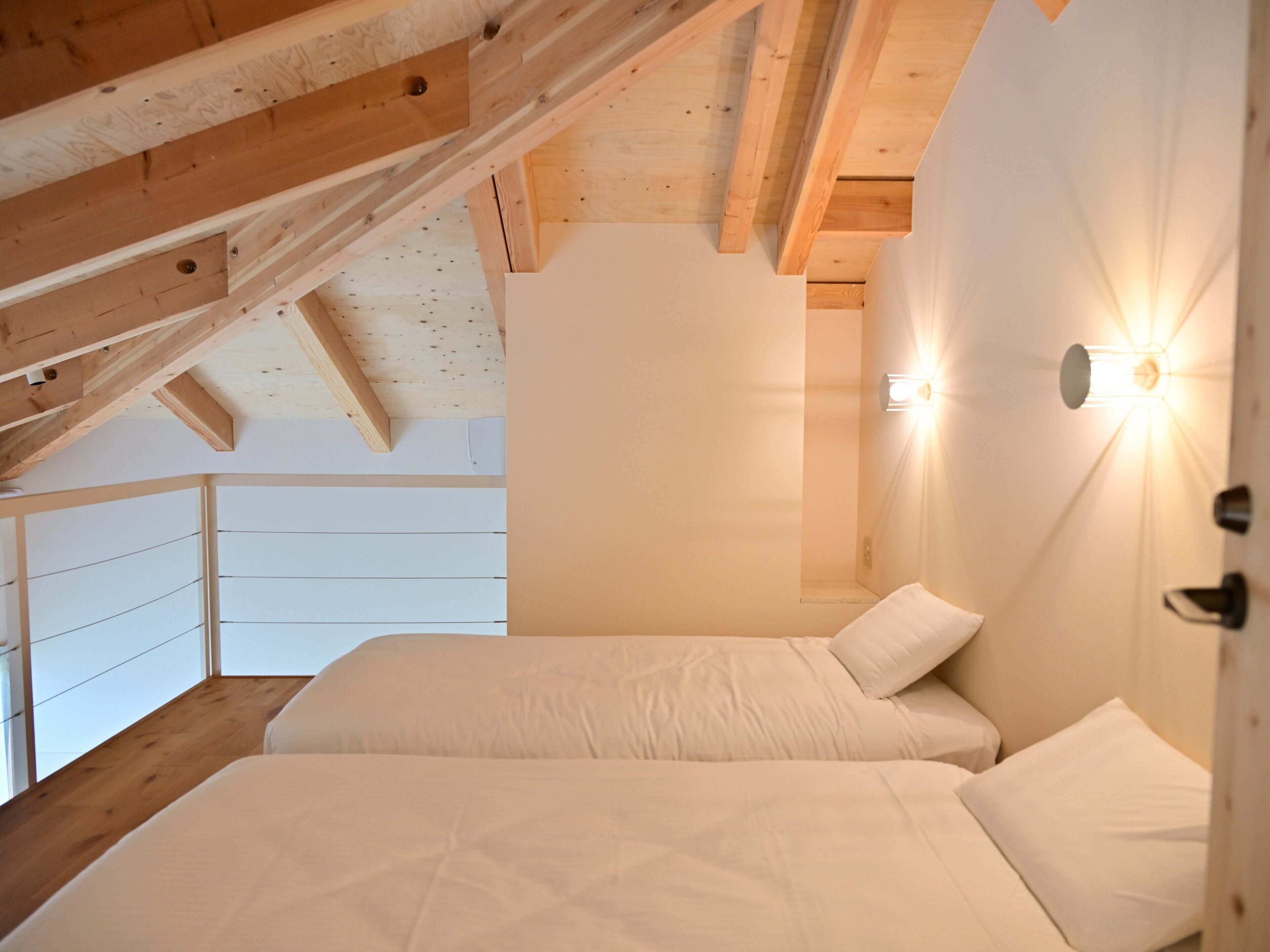 Quarto com teto inclinado, vigas de madeira e duas camas com roupa de cama branca.