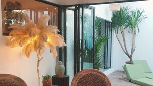 Uma área de jantar com mesa de madeira, cadeiras, uma escultura decorativa de pássaro, um abajur em formato de palmeira e vista para o exterior através de grandes portas de vidro.