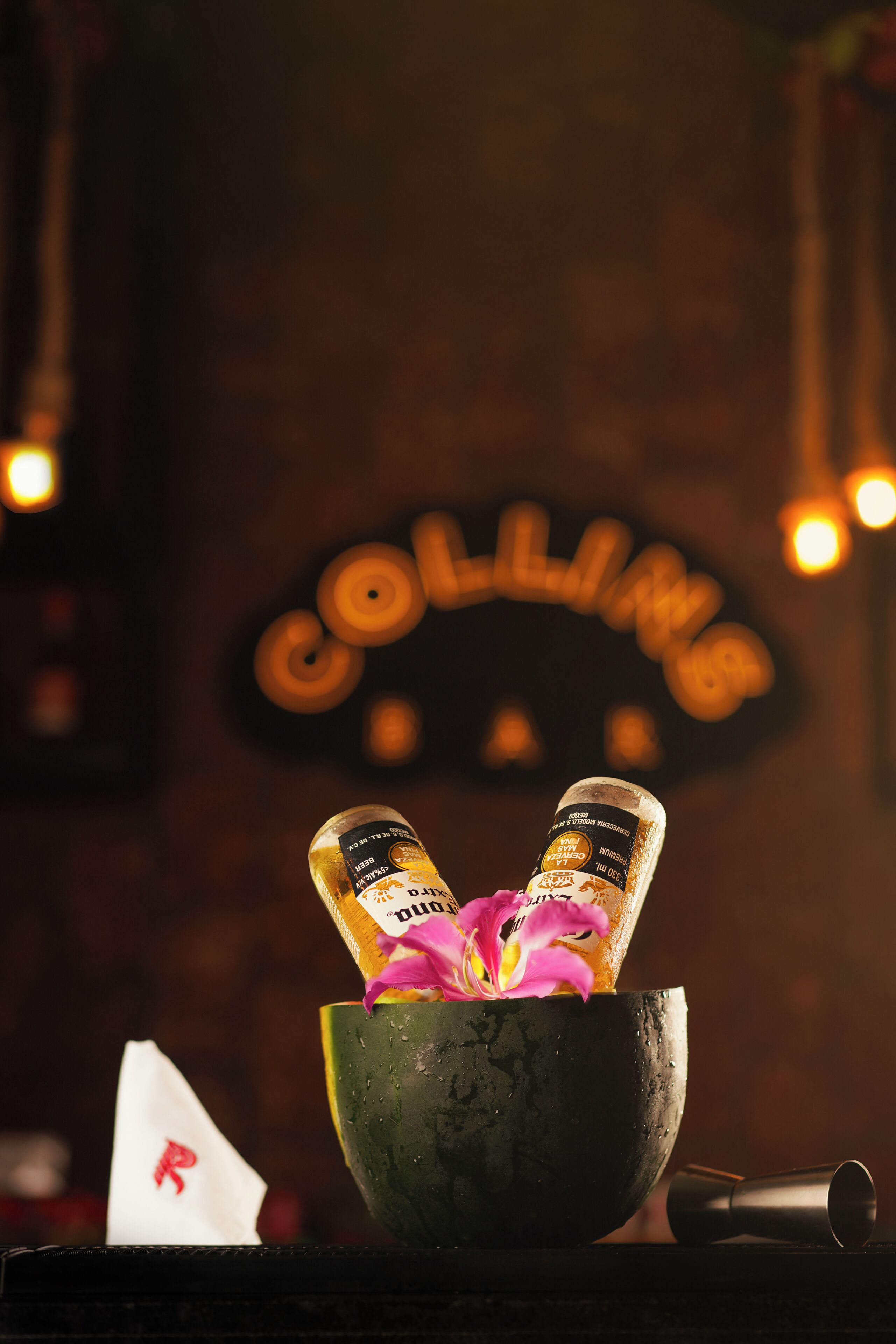 Ambiente rústico com um letreiro de neon que diz "Collins" ao fundo, uma tigela verde com duas garrafas e flores cor-de-rosa, e um guardanapo branco com um logotipo vermelho.