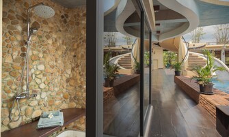Compartimento de chuveiro em pedra, um banco de madeira com toalhas, e área de piscina com plantas em vasos.