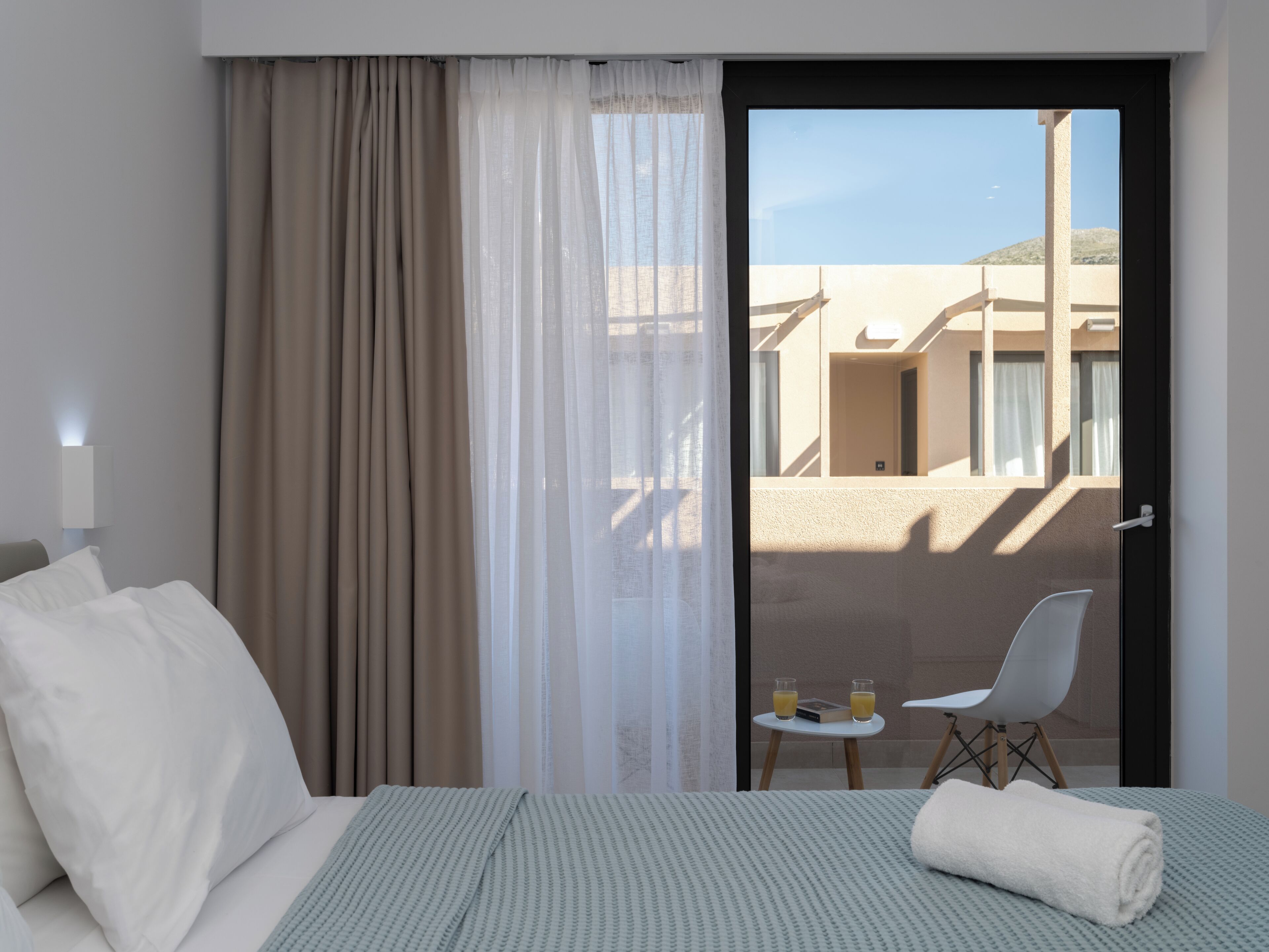 Quarto de hotel com uma cama, uma mesinha com dois copos de suco de laranja, uma cadeira branca e vista para um edifício através de uma porta de vidro.
