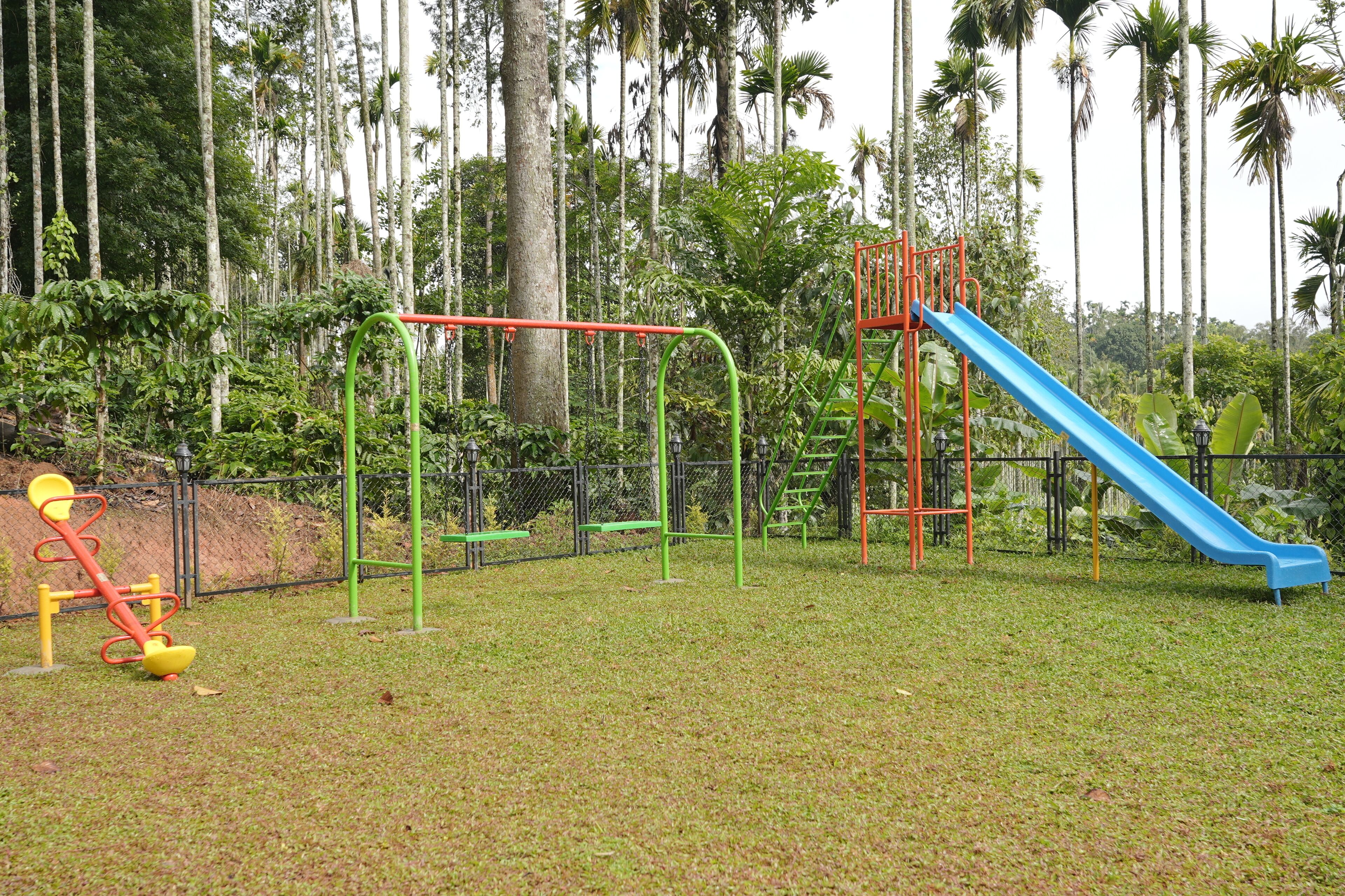 Um parquinho com um escorrega azul, estruturas de escalada vermelhas e verdes e um balanço amarelo.