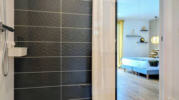 Um banheiro moderno com chuveiro, paredes de azulejo escuro, uma pia branca e uma porta de vidro que dá para um quarto com cama e mesa de cabeceira.