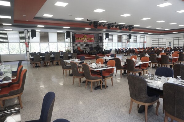 Uma ampla área de refeições de hotel, com várias mesas e cadeiras, um palco com instrumentos musicais e grandes janelas que permitem entrada de luz natural.