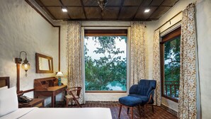 Quarto de hotel com uma cama grande, uma escrivaninha com luminária, uma cadeira e vista para árvores pela janela.