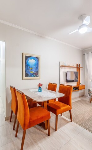Uma sala de jantar com mesa branca, cadeiras laranja, um quadro emoldurado e uma televisão sobre um suporte de madeira.