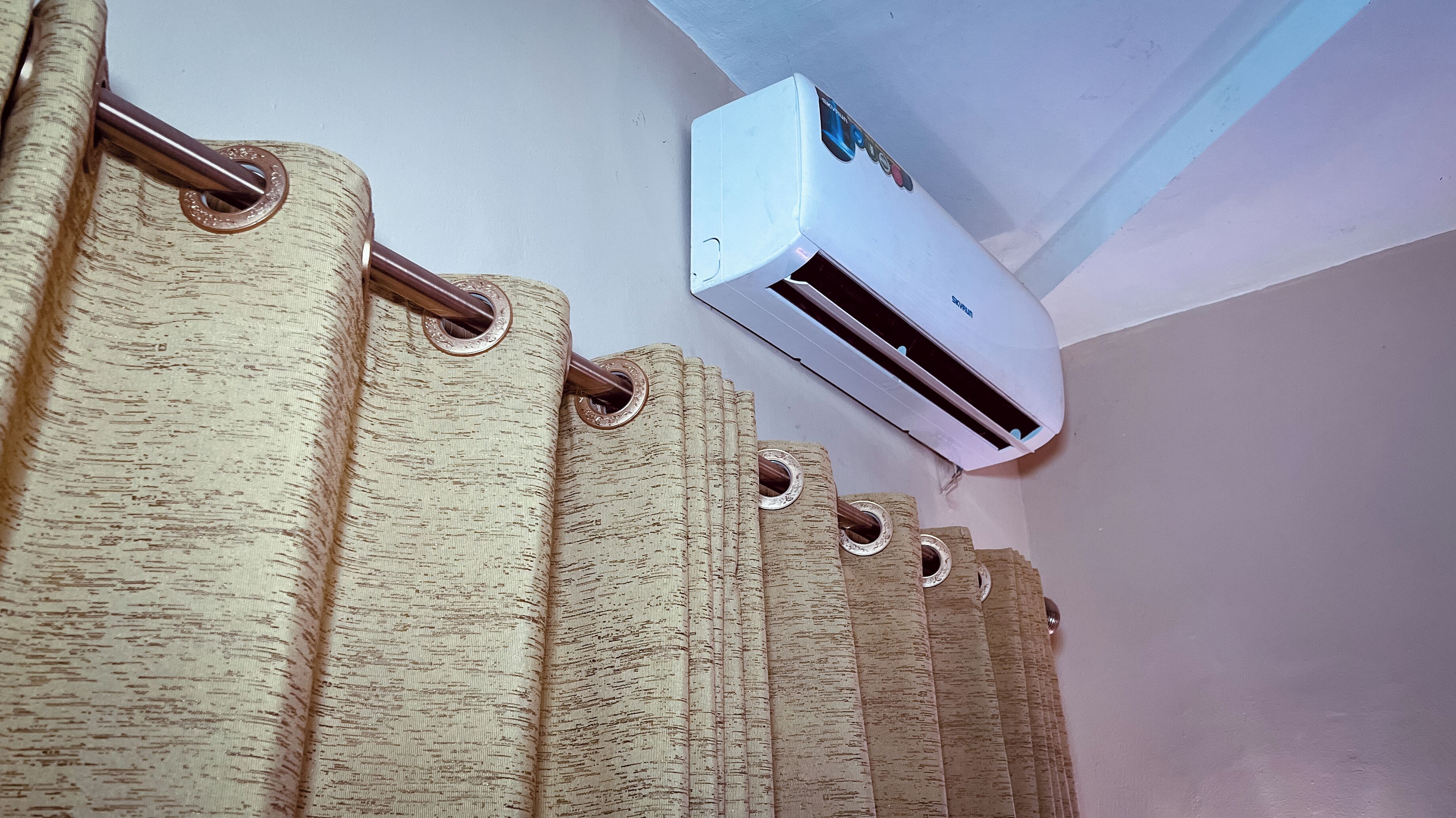 Unidade de ar condicionado suspensa no teto acima de cortinas bege com vários furos.