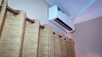 Unidade de ar condicionado suspensa no teto acima de cortinas bege com vários furos.