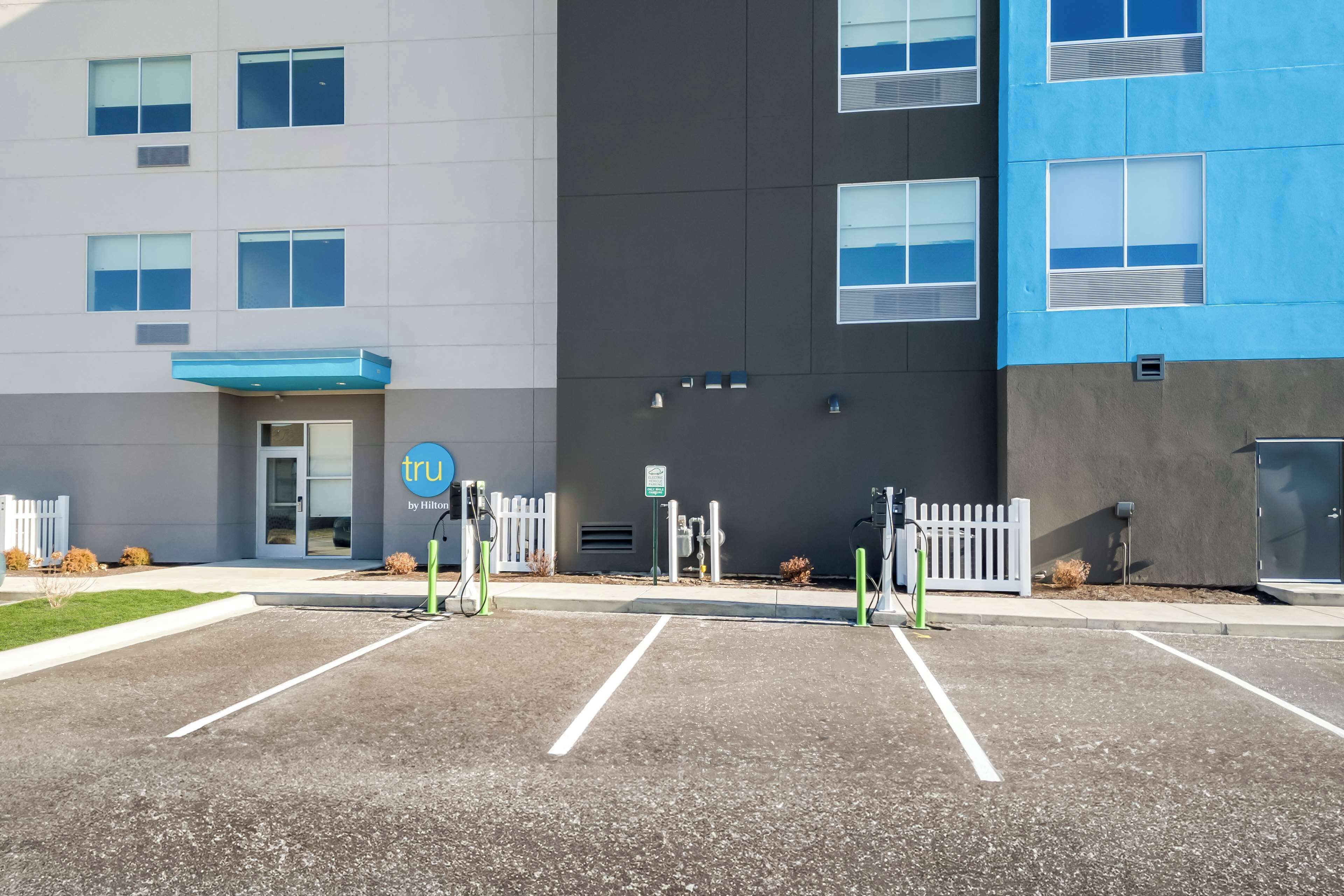 Um estacionamento com estações de carregamento para veículos elétricos, um edifício com fachada azul e cinza, e uma cerca branca de piquete.