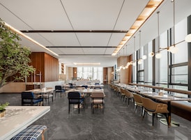 Restaurante moderno com área de refeições extensa, várias mesas e cadeiras. Grandes janelas proporcionam luz natural e vista da cidade.