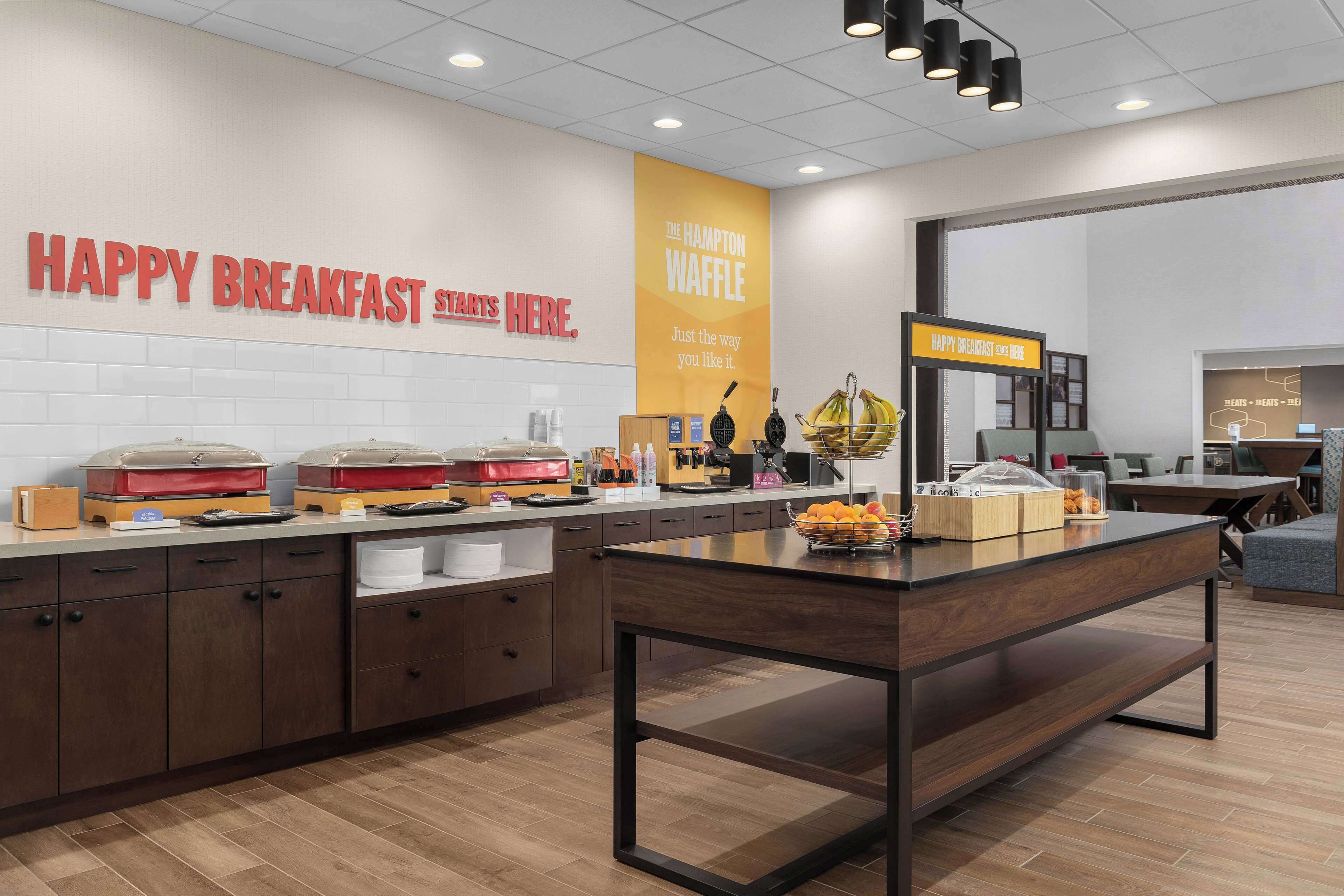 Uma área de café da manhã com um balcão exibindo alimentos, uma parede com a placa 'HAPPY BREAKFAST STARTS HERE' e a placa 'THE HAMPTON WAFFLE'.