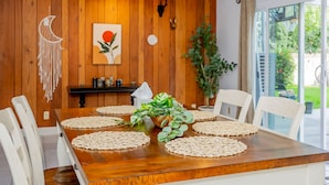 Uma mesa de jantar de madeira com jogos americanos, cadeiras brancas, uma planta em vaso e uma obra de arte pendurada na parede.