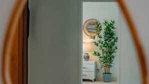 Corredor com planta em vaso, luminária e espelho refletindo um quarto com cama e cômoda.
